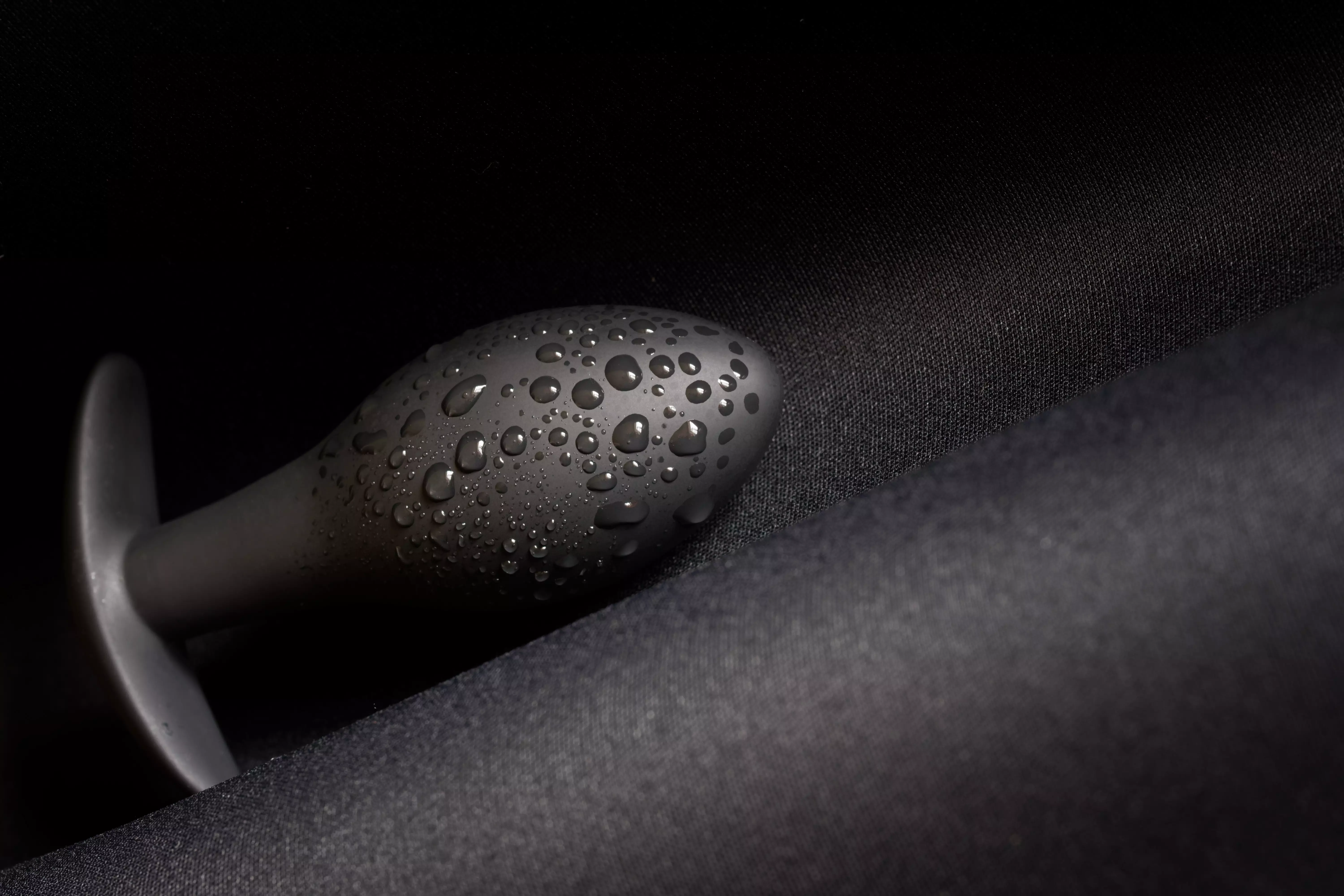 Een natte zwarte buttplug ligt op een donkere ondergrond. Vocht- of smeermiddeldruppels. Seksspeeltje en plezier. Diagonaal