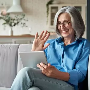 60 च्या दशकातील आनंदी प्रौढ मध्यमवयीन प्रौढ स्त्री हातात हात हलवत डिजिटल टॅबलेट संगणक व्हिडिओ कॉन्फरन्स कॉलिंग सोशल डिस्टन्स व्हर्च्युअल फॅमिली ऑनलाइन चॅट मीटिंग घरी सोफ्यावर बसून.