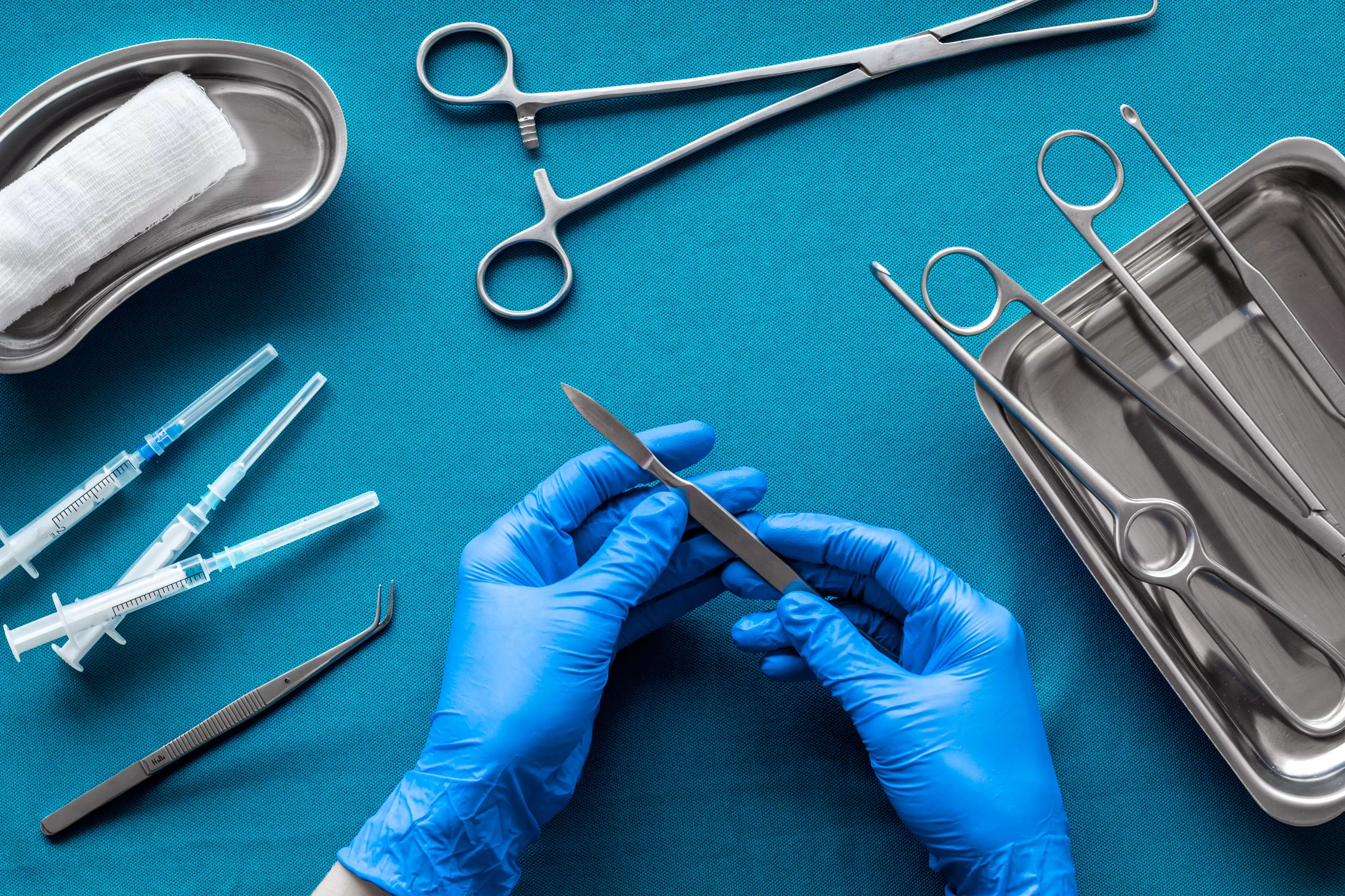 Përgatitja për kirurgji plastike. Duart e mjekut marrin bisturi në sfond blu me pamje të sipërme të mjeteve kirurgjikale.